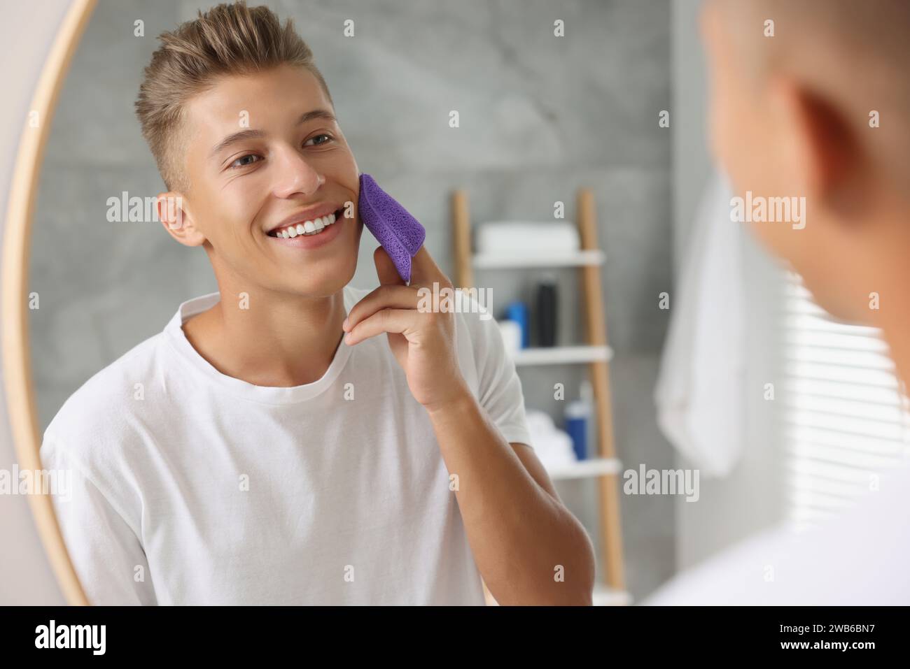 Happy young man washing his face with sponge near mirror in bathroom ...