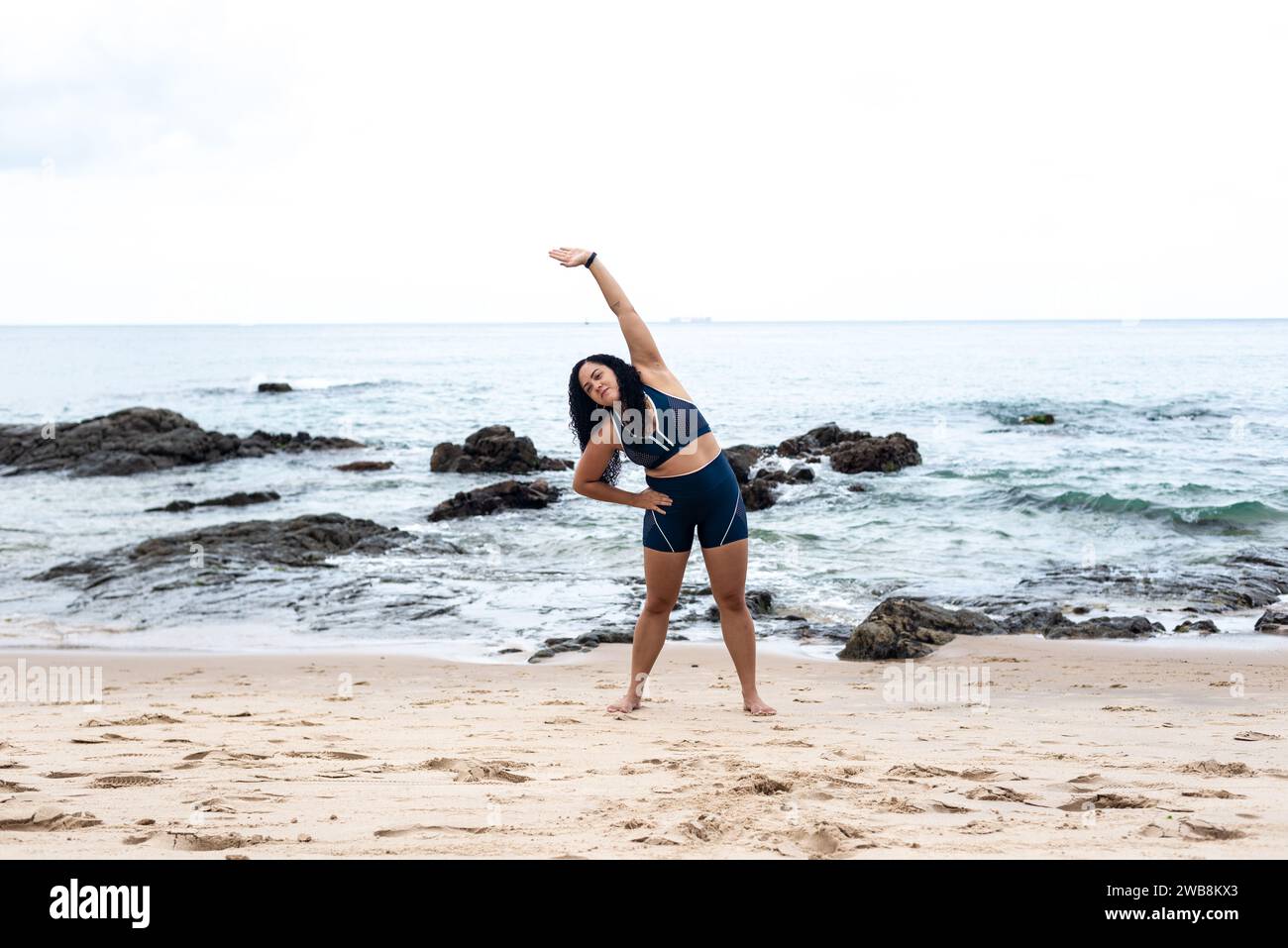 Beautiful young woman in gym clothes doing stretches on the beach sand ...