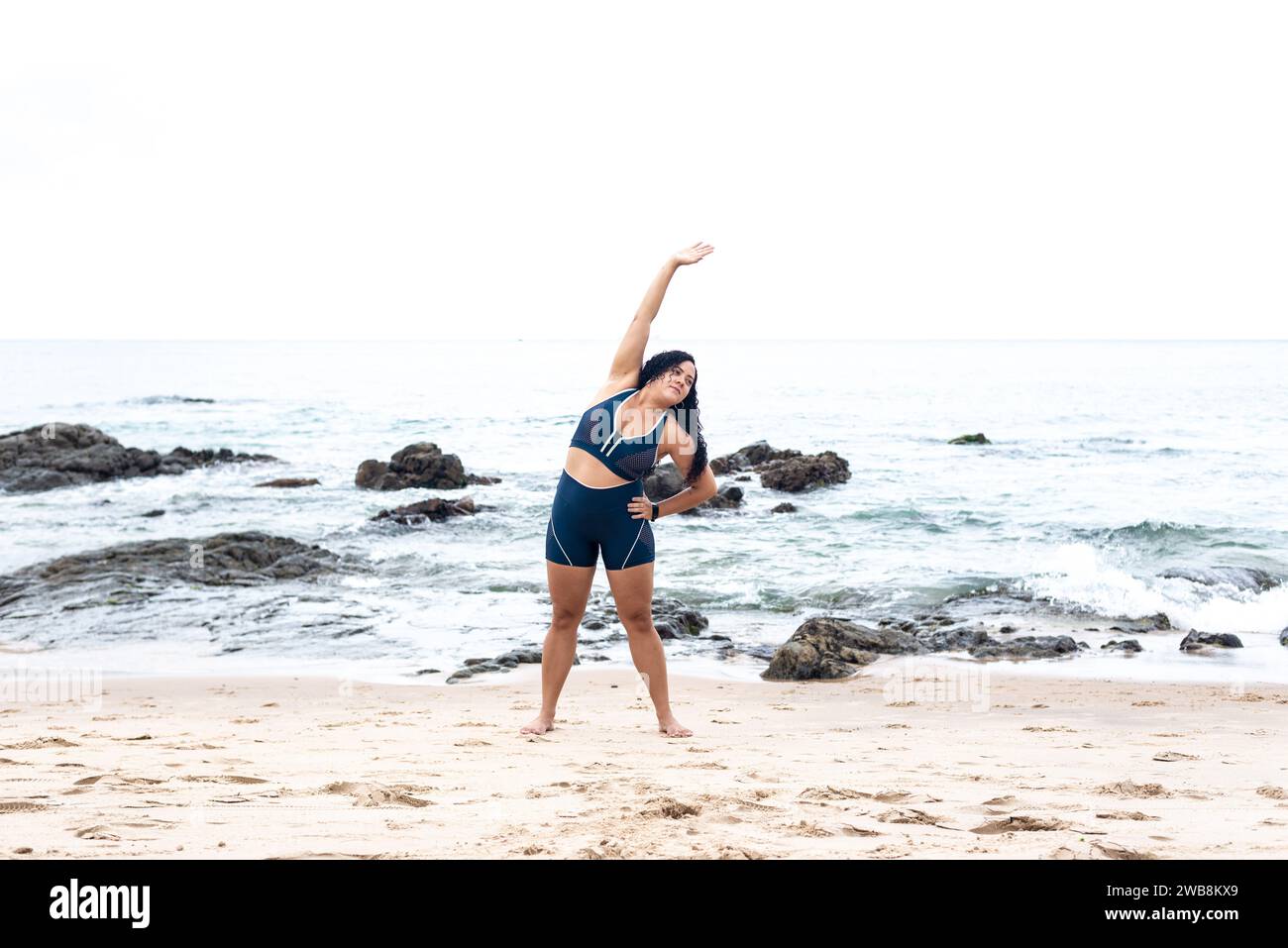 Beautiful young woman in gym clothes doing stretches on the beach sand ...