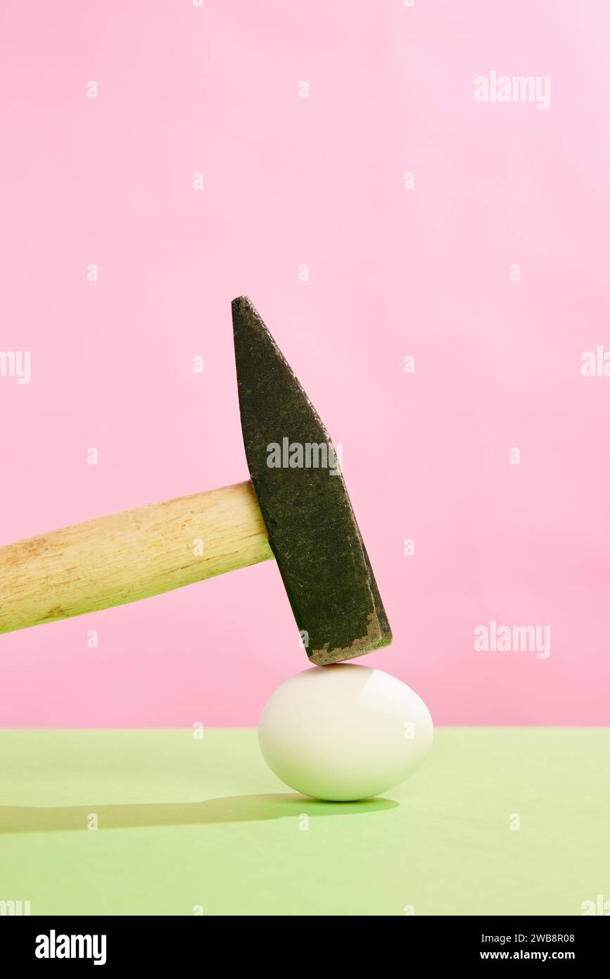 Hammer cracking white egg against pink studio background. Ester ...