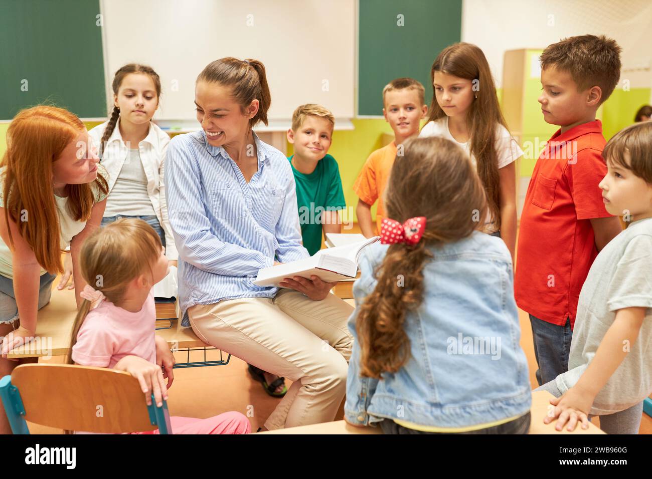 Happy female teacher reading book to schoolkids while sitting on bench ...