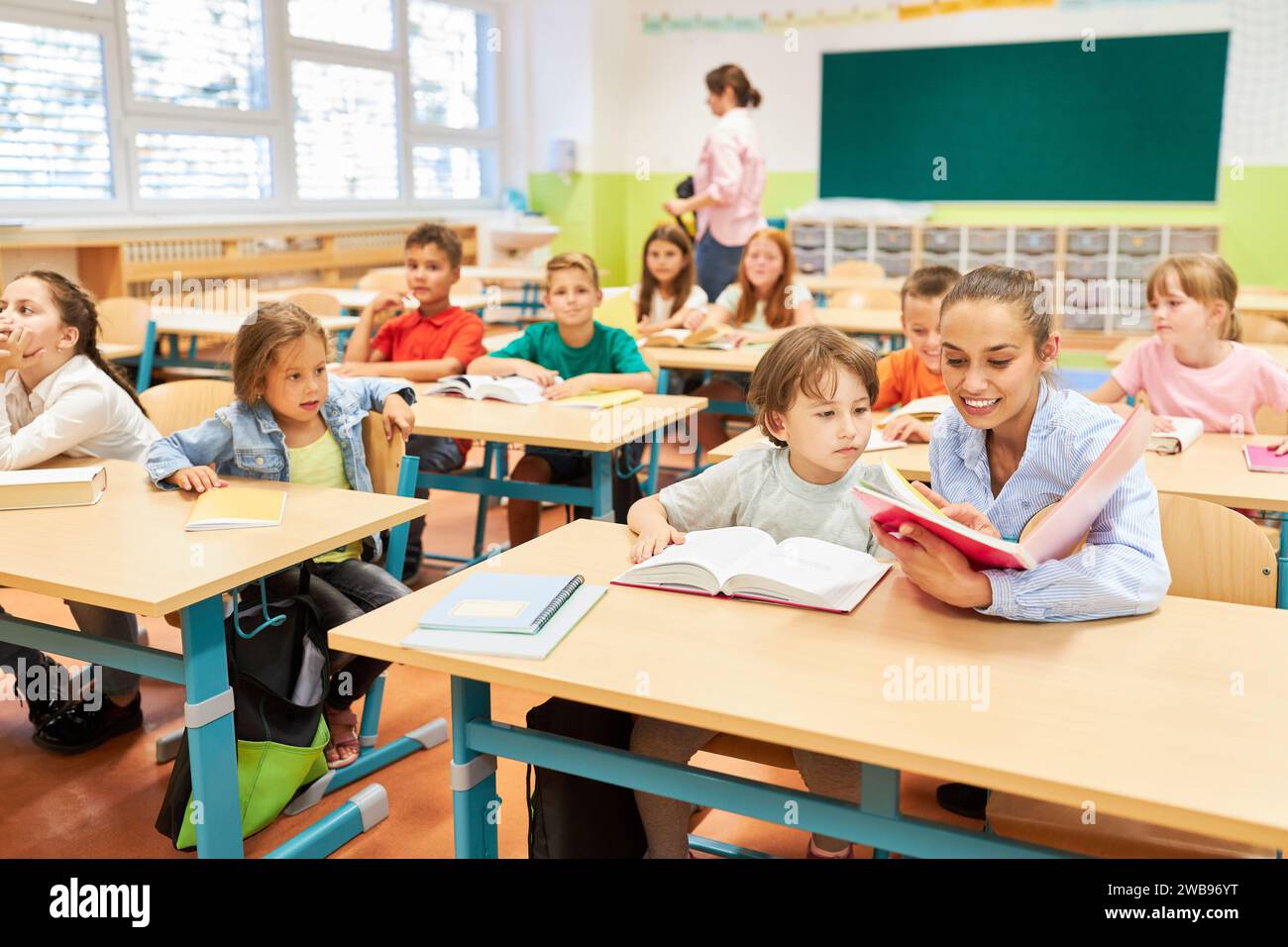 Smiling elementary teacher reading book to schoolgirl in classroom ...