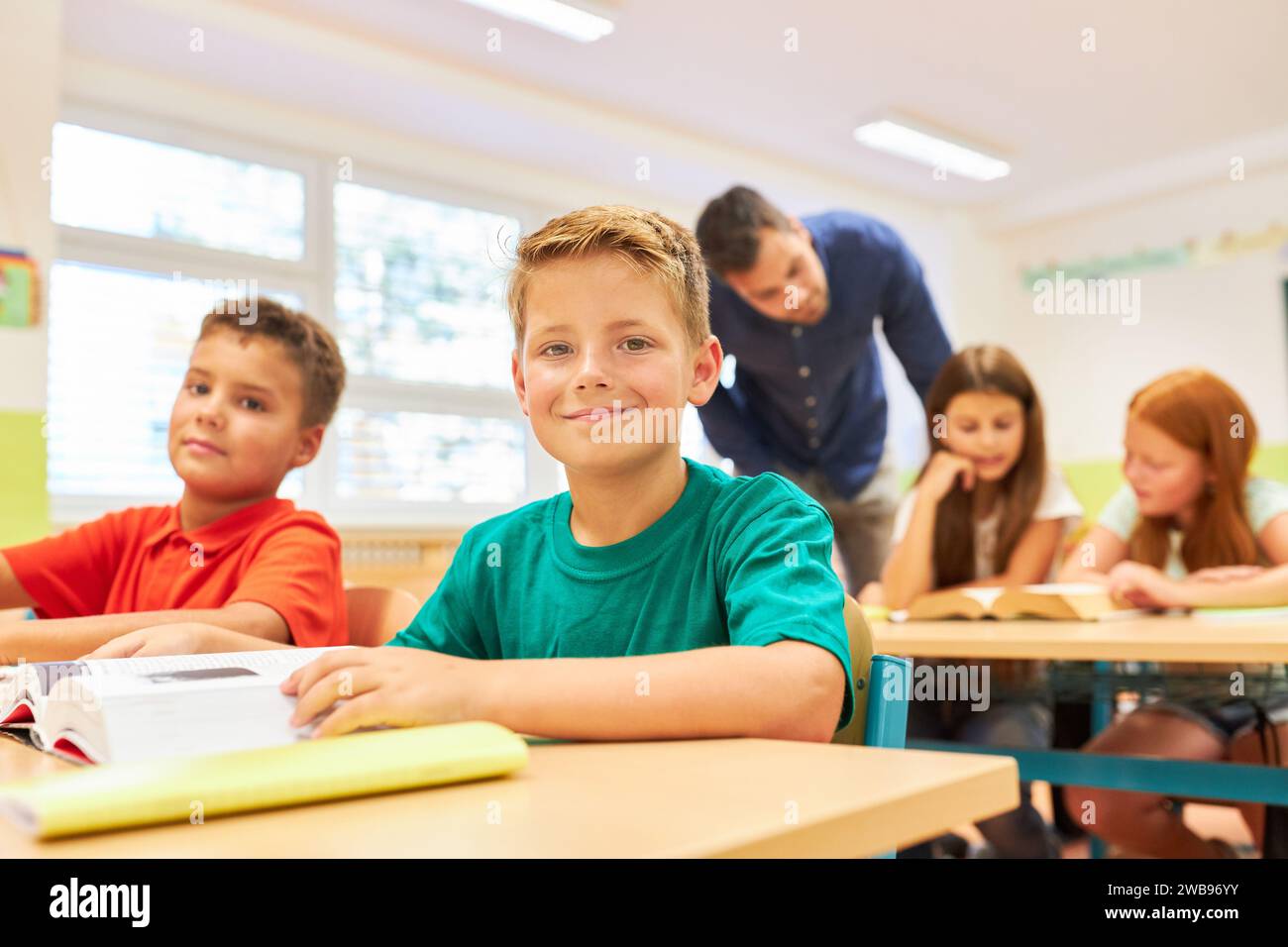 Portrait of smiling schoolboy sitting with book on bench in classroom ...