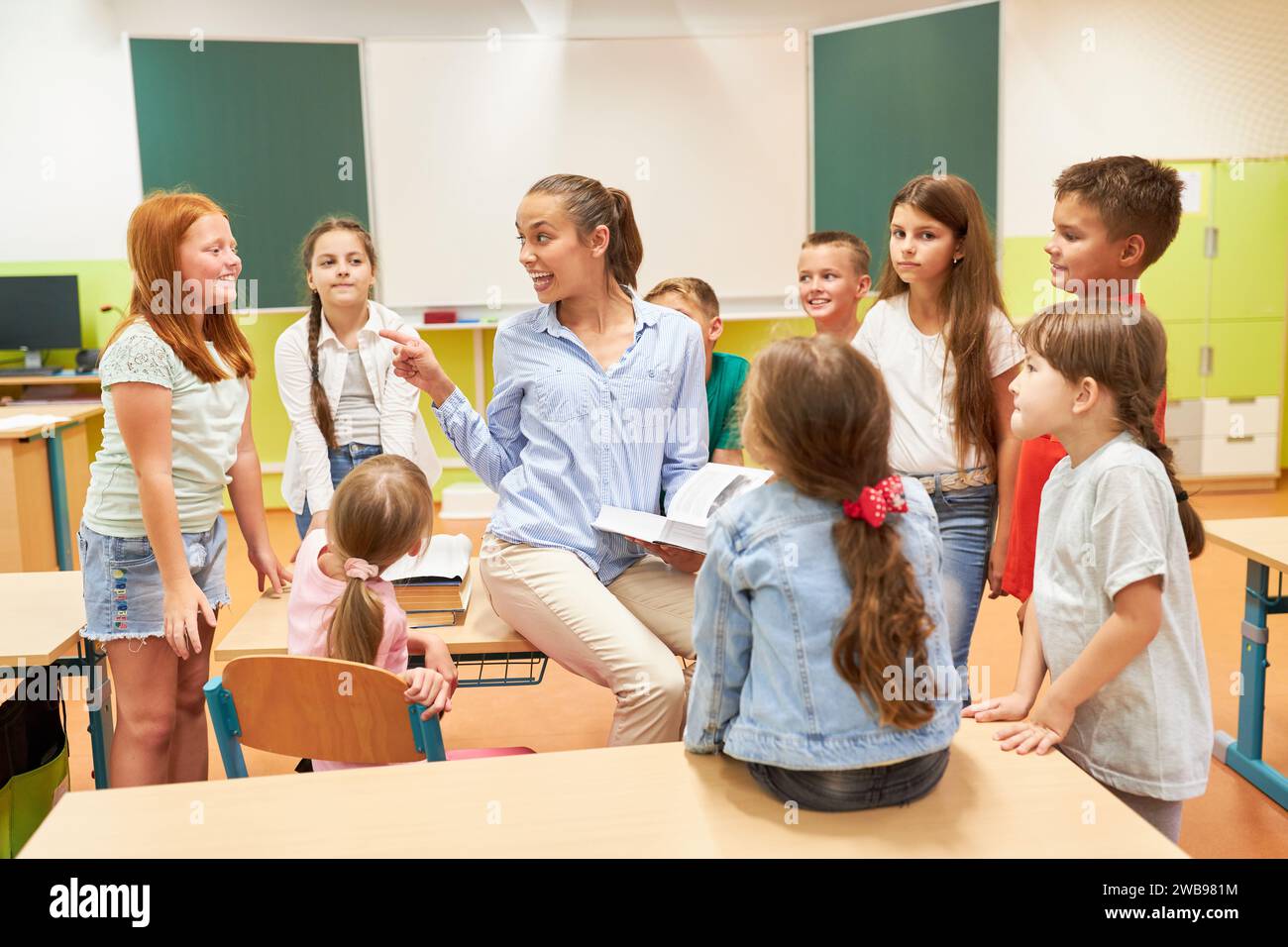 Happy female teacher pointing at girl while sitting amidst students in ...
