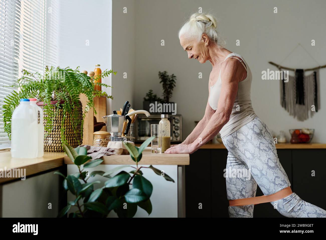 Active aged woman with resistance band around legs standing by kitchen ...