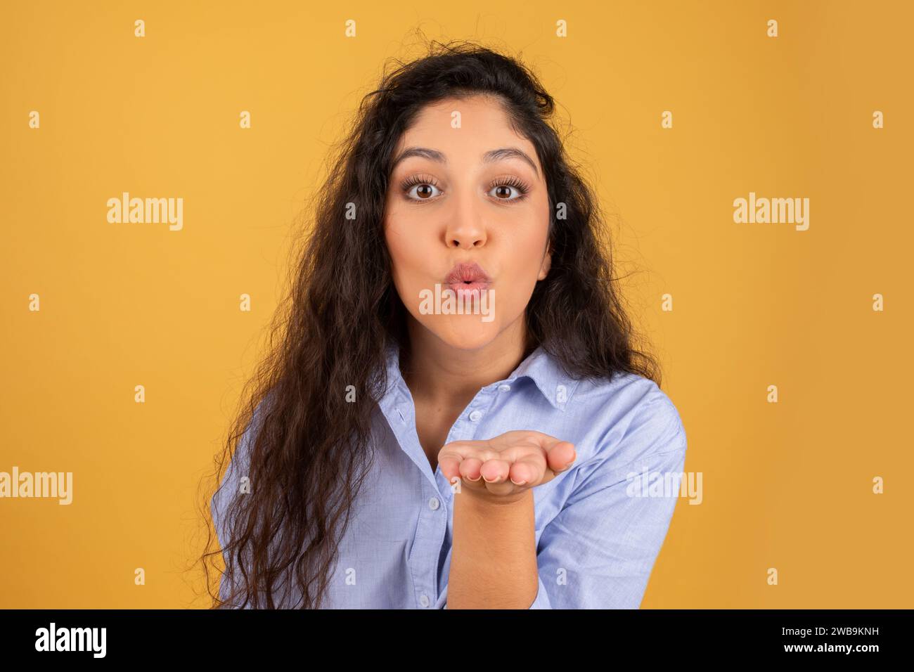 A young woman with long wavy hair, wearing a light blue shirt, blows a ...