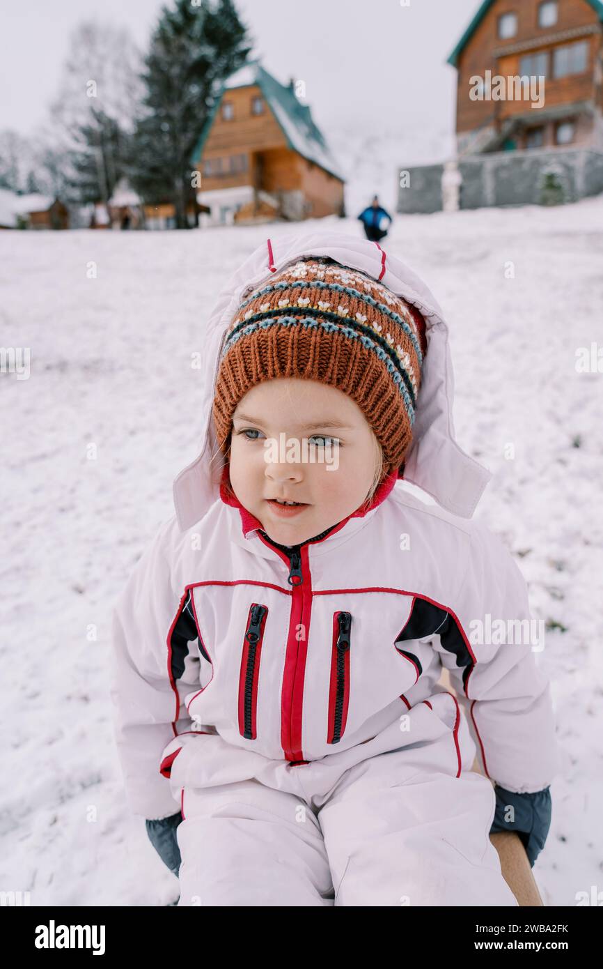 Little smiling girl sits on a sled at the bottom of a snowy hill and ...