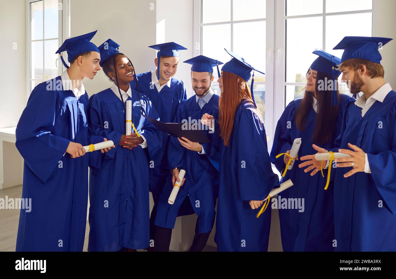 Happy graduate students in blue graduation robes standing indoors with ...