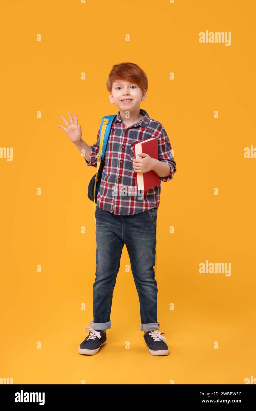 Happy schoolboy with backpack and book waving hello on orange ...