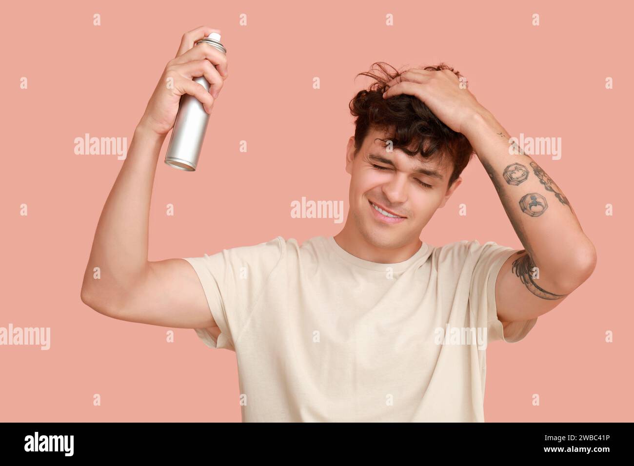 Handsome young man applying hair spray on his curly hair against pink ...