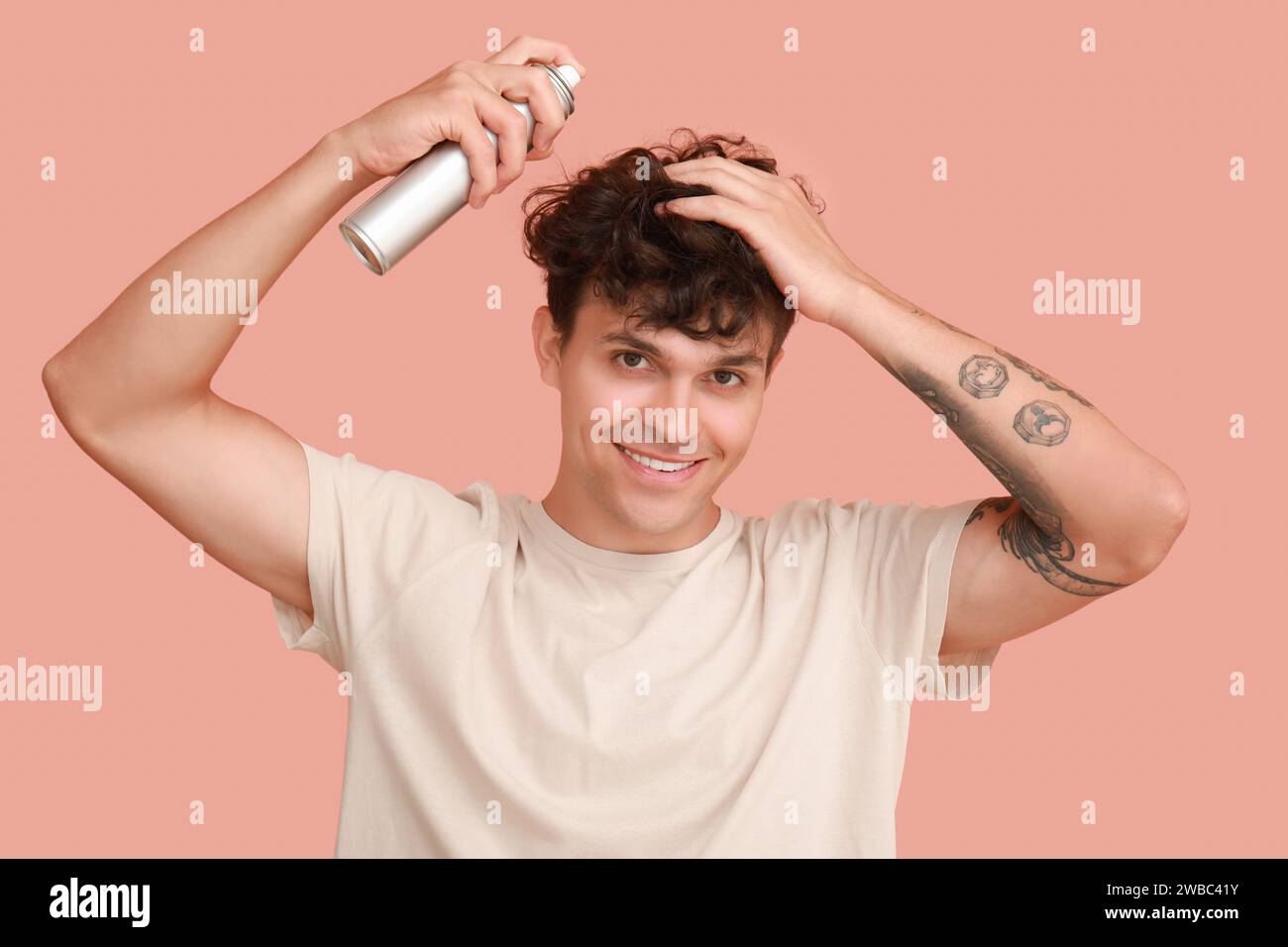 Handsome young man applying hair spray on his curly hair against pink ...
