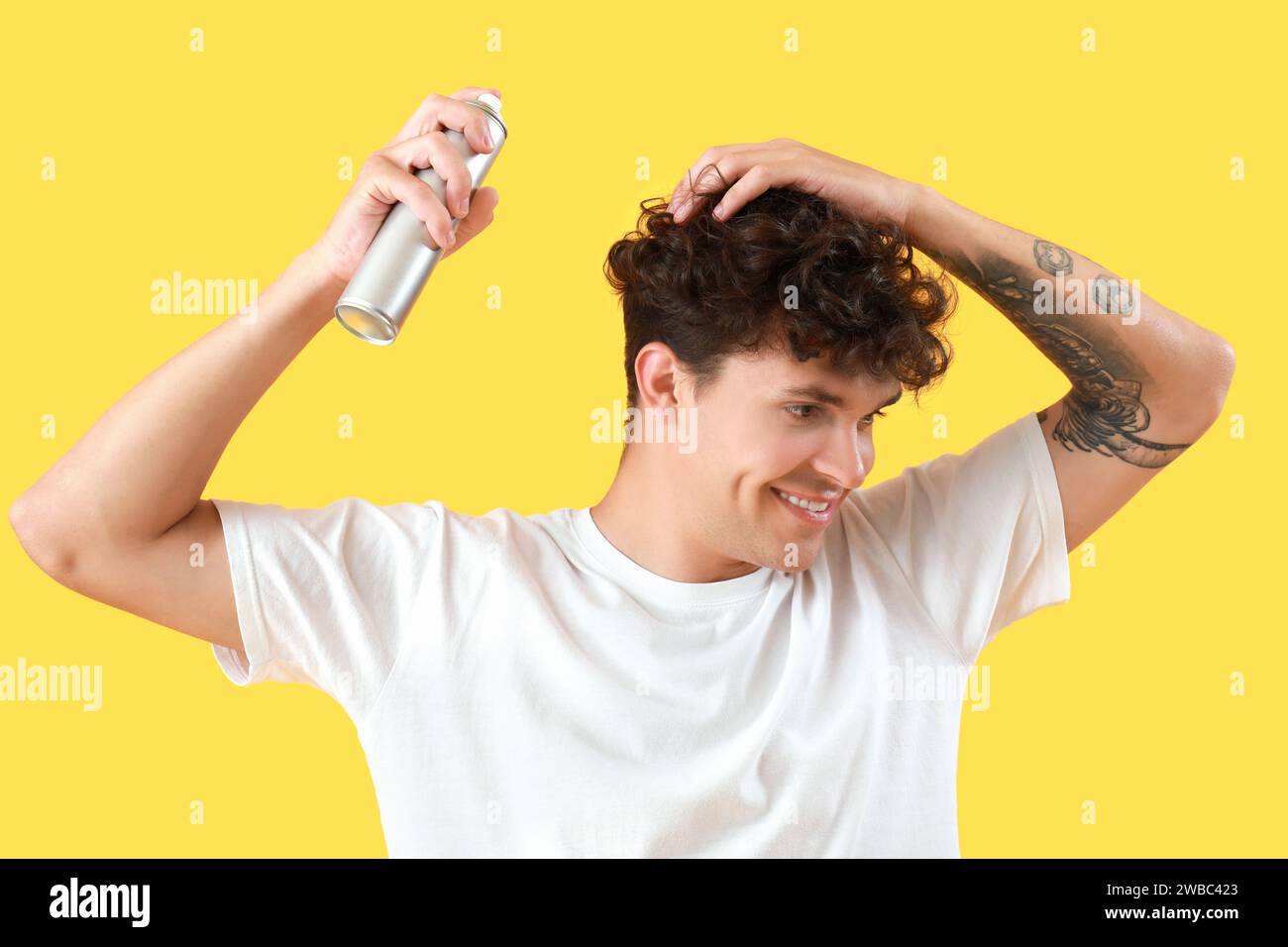 Handsome young man applying hair spray on his curly hair against yellow ...