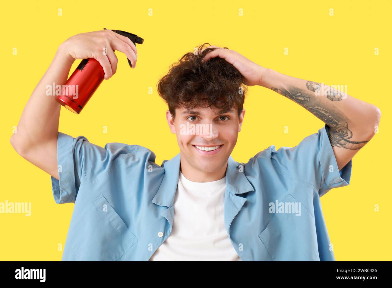 Handsome young man applying hair spray on his curly hair against yellow ...