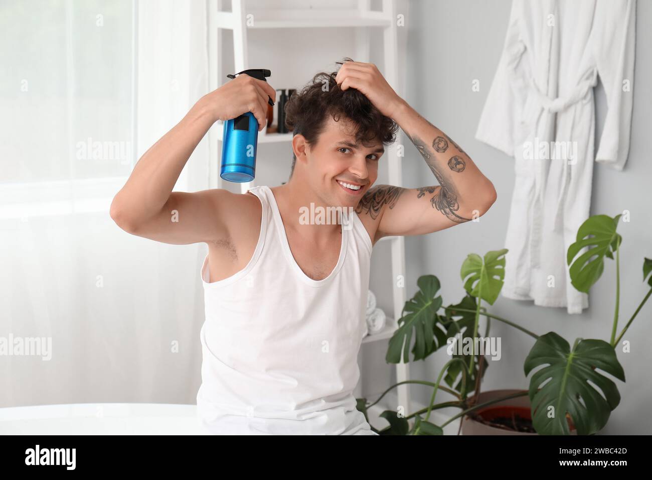 Handsome young man with tattoo applying hair spray on his curly hair in ...