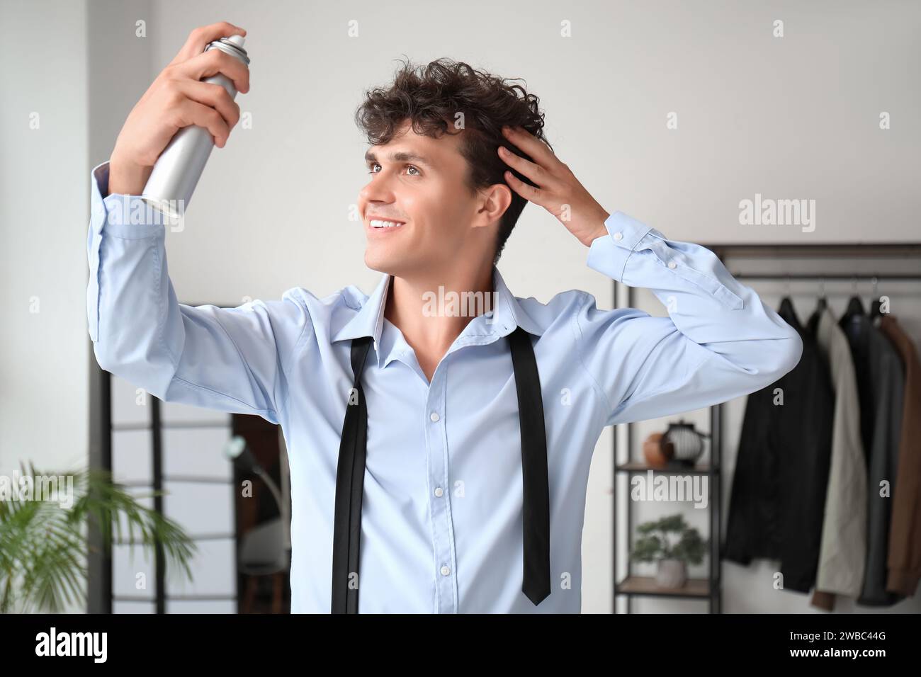 Handsome young man applying hair spray on his curly hair in hallway ...
