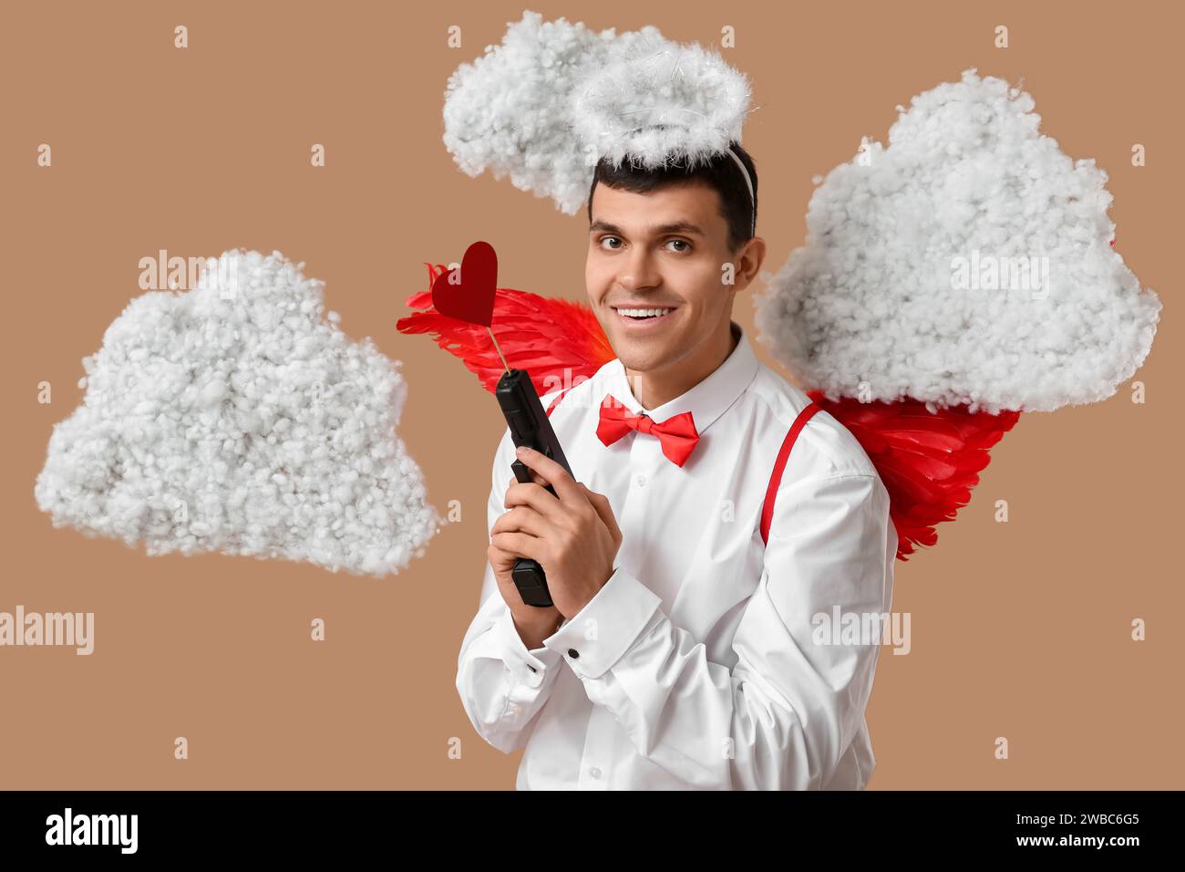 Cool young man dressed as Cupid with gun in clouds on beige background ...