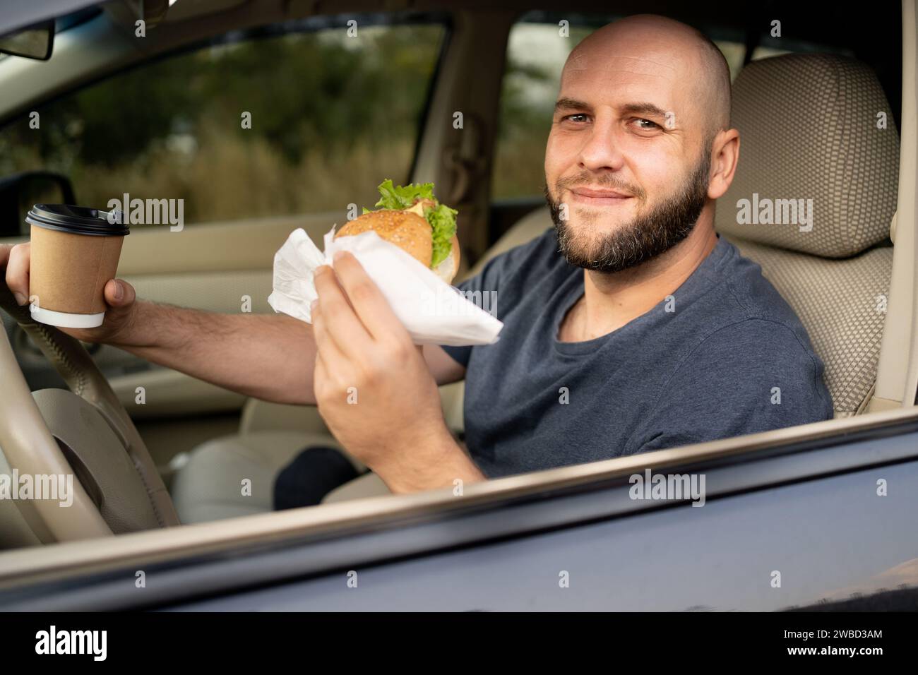 A bald smiling man driving a car drinking takeaway coffee and eating a ...