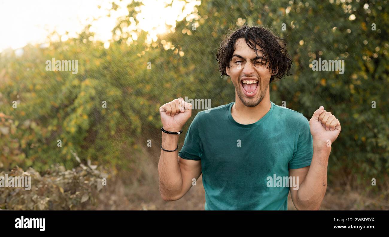 Smiling young man during the rain in the park. Guy enjoying the rain ...