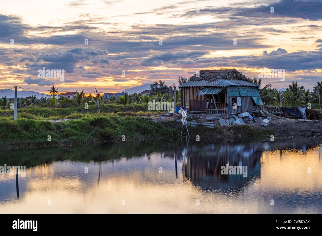 An old shack, located by a river in central Vietnam. The sun is setting through a cloudy sky ...