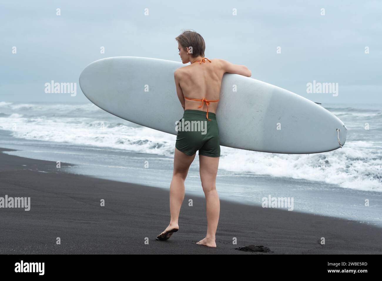 Rear view of female surfer walking on black sandy beach carrying white ...