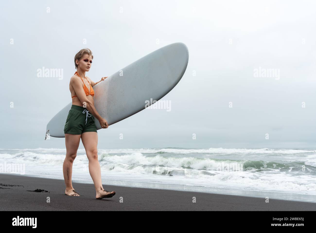 Woman surfer walks on black sand beach carrying white surfboard against ...