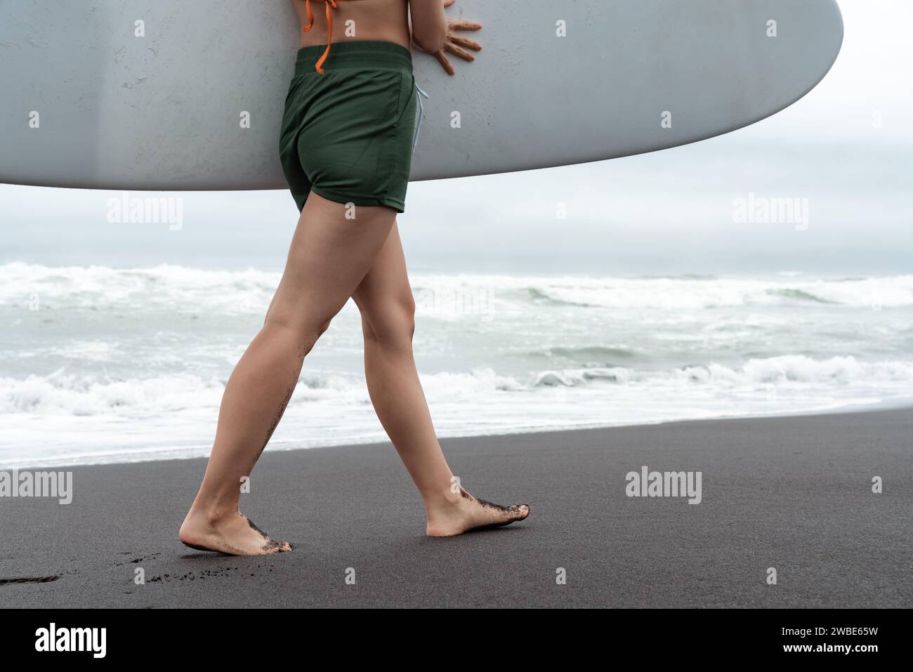 Surfer woman carrying surfboard walking along beach with sea waves in ...