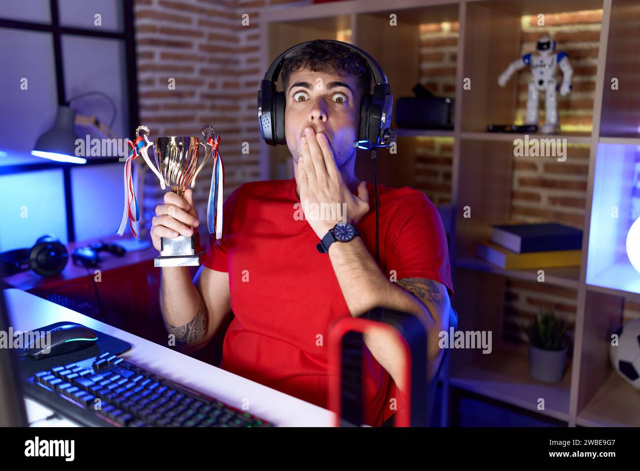 Young hispanic man playing video games holding trophy covering mouth ...