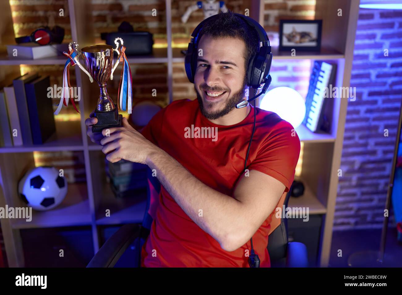 Young hispanic man playing video games holding trophy looking positive ...