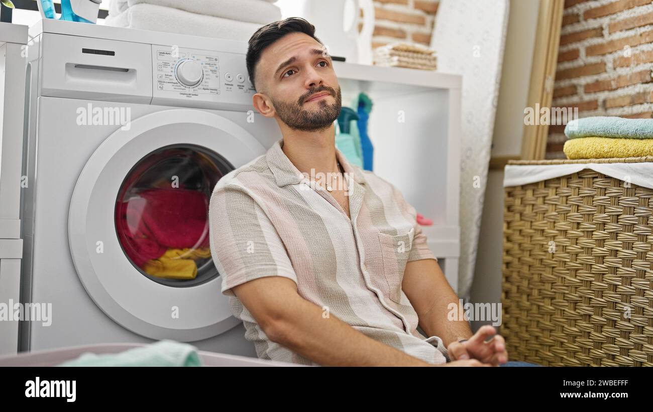 Young hispanic man leaning on washing machine stressed at laundry room ...