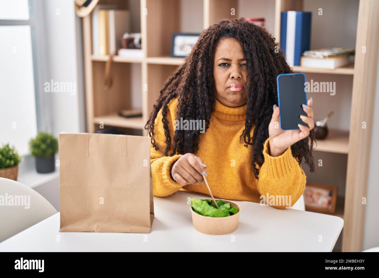 Plus size hispanic woman eating take away salad holding smartphone ...