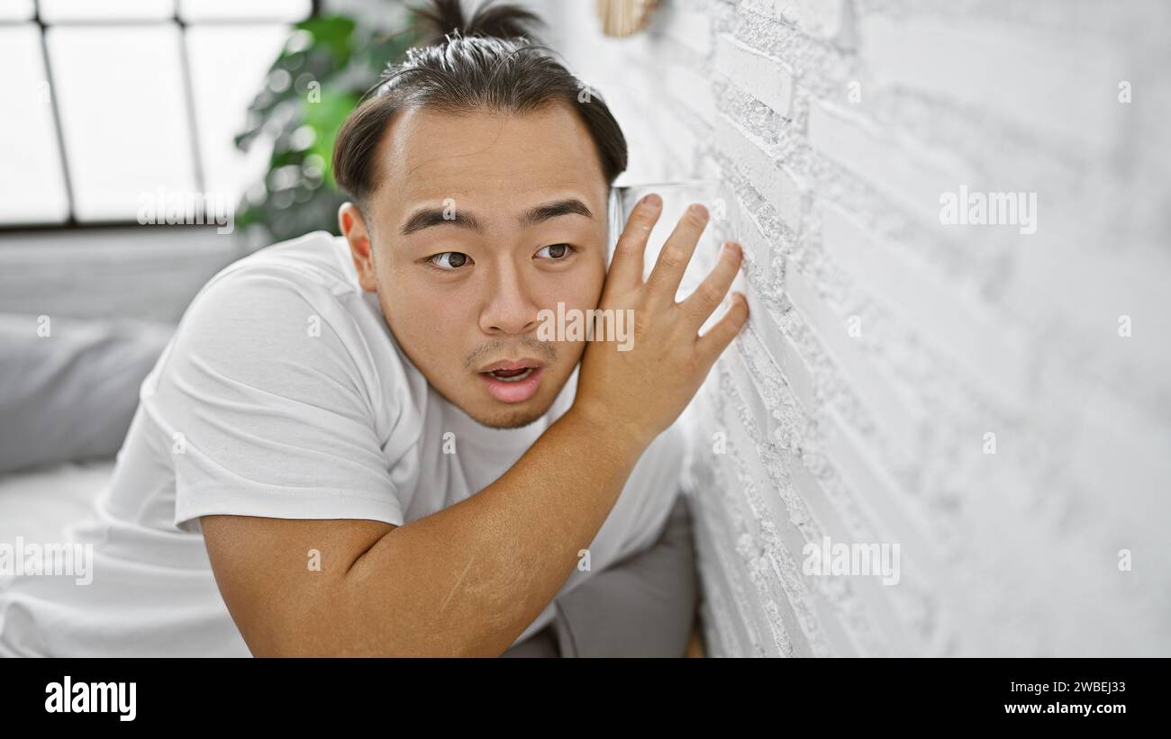 Nosy young chinese man sneakily listening through bedroom wall with glass, overcome by curiosity ...
