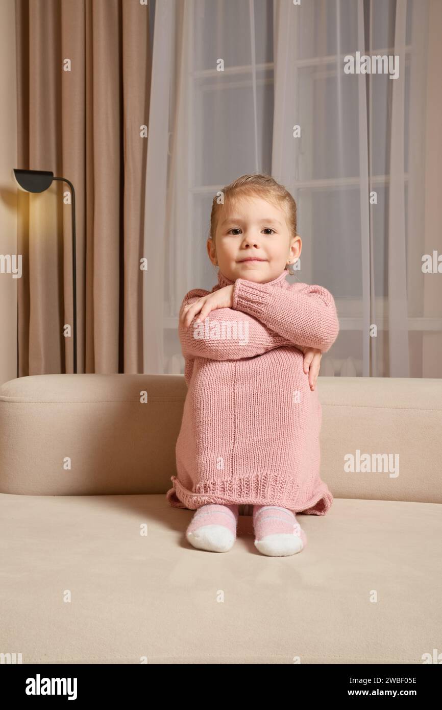Cute little girl sitting on the sofa with her pink sweater pulled over ...