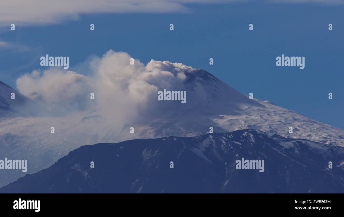 Mount Etna. Smoke rises from a snow-covered volcano under a clear blue ...