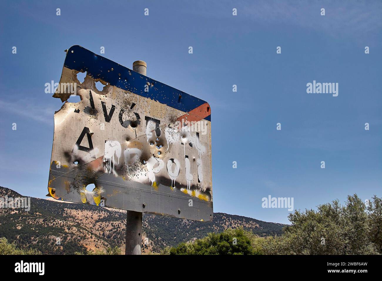 Weathered road sign with bullet holes against a blue sky and trees in ...
