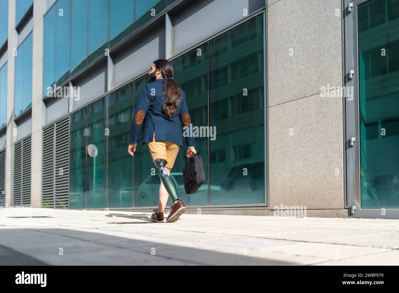 Rear view of a young businessman with amputee leg walking along a ...