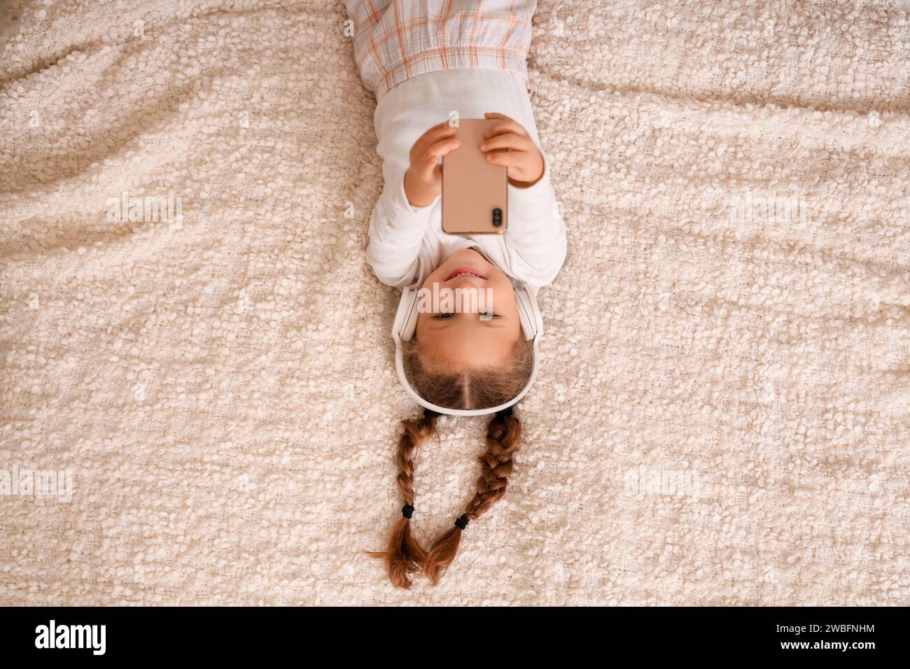 Cute little girl in headphones with mobile phone lying on bed, top view ...