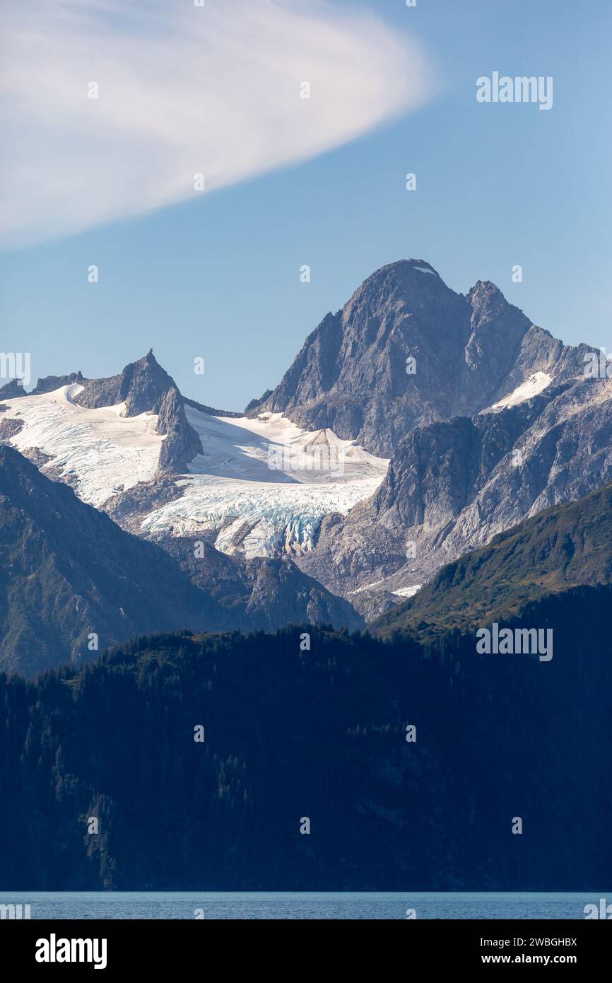 Glacier flowing down a jagged mountain peaks rise above calm blue ...