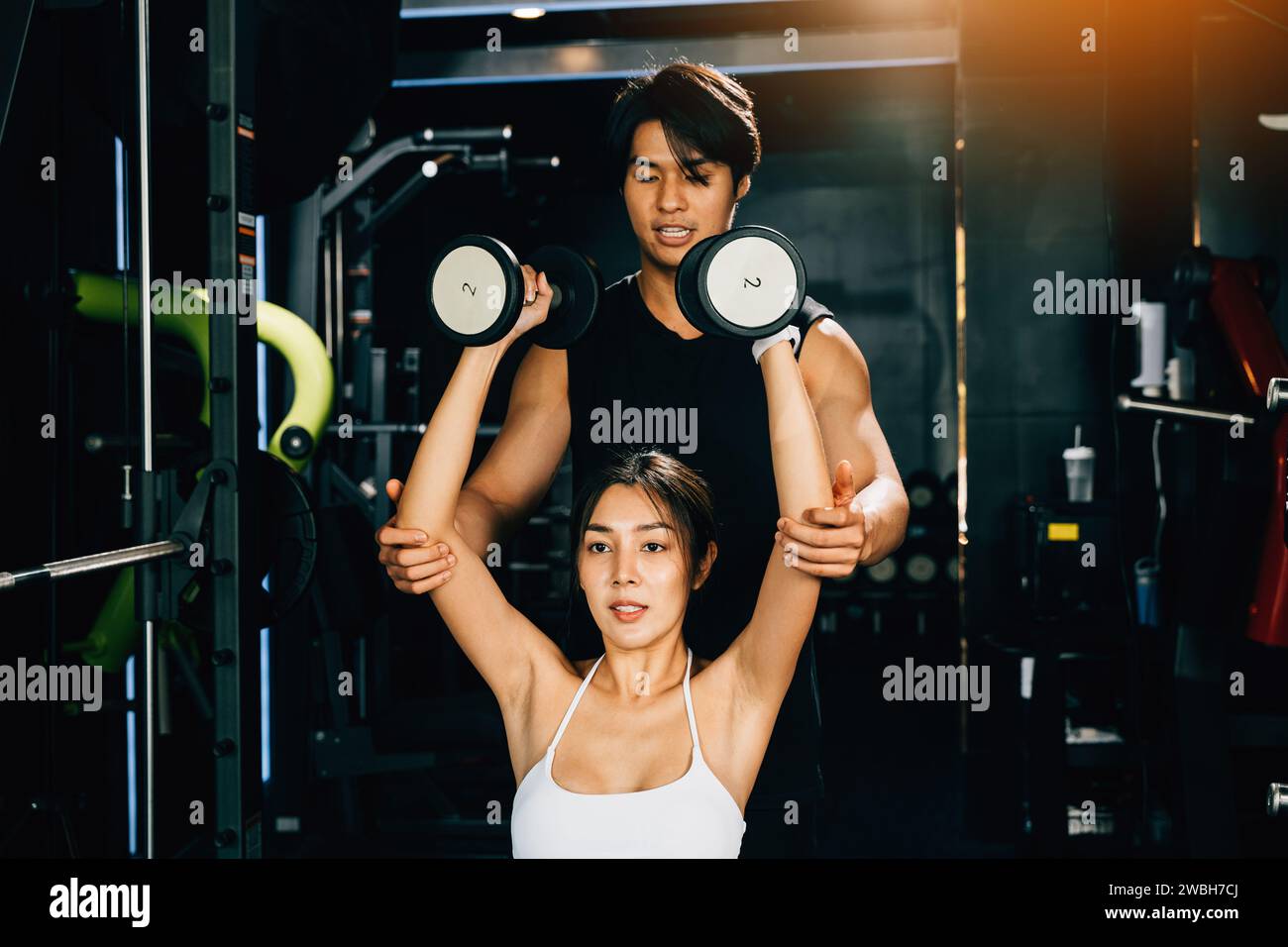 Fit Asian woman lifting dumbbells with a smile, supported by her ...