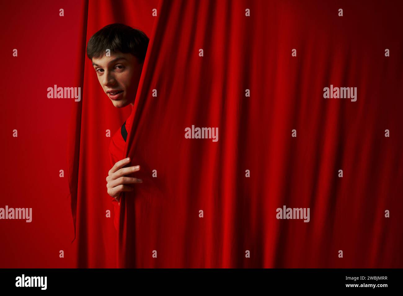 playful young man in vibrant shirt hiding behind red curtain while ...