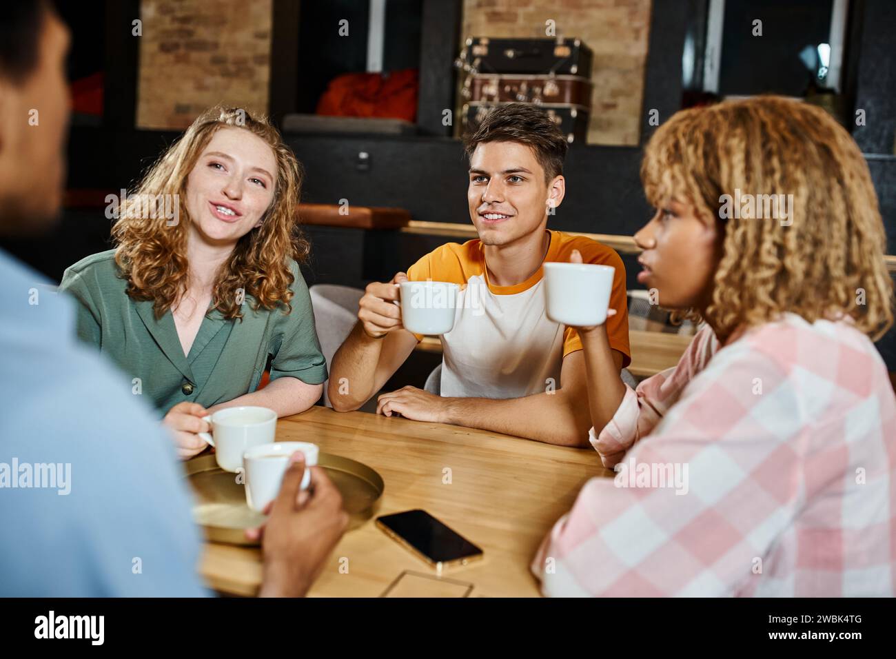 positive multiethnic friends with coffee cups talking in lounge cafe of ...