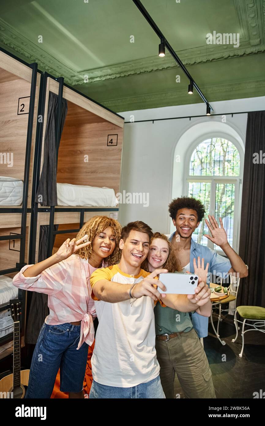 carefree multiethnic students waving hands and taking selfie in modern ...