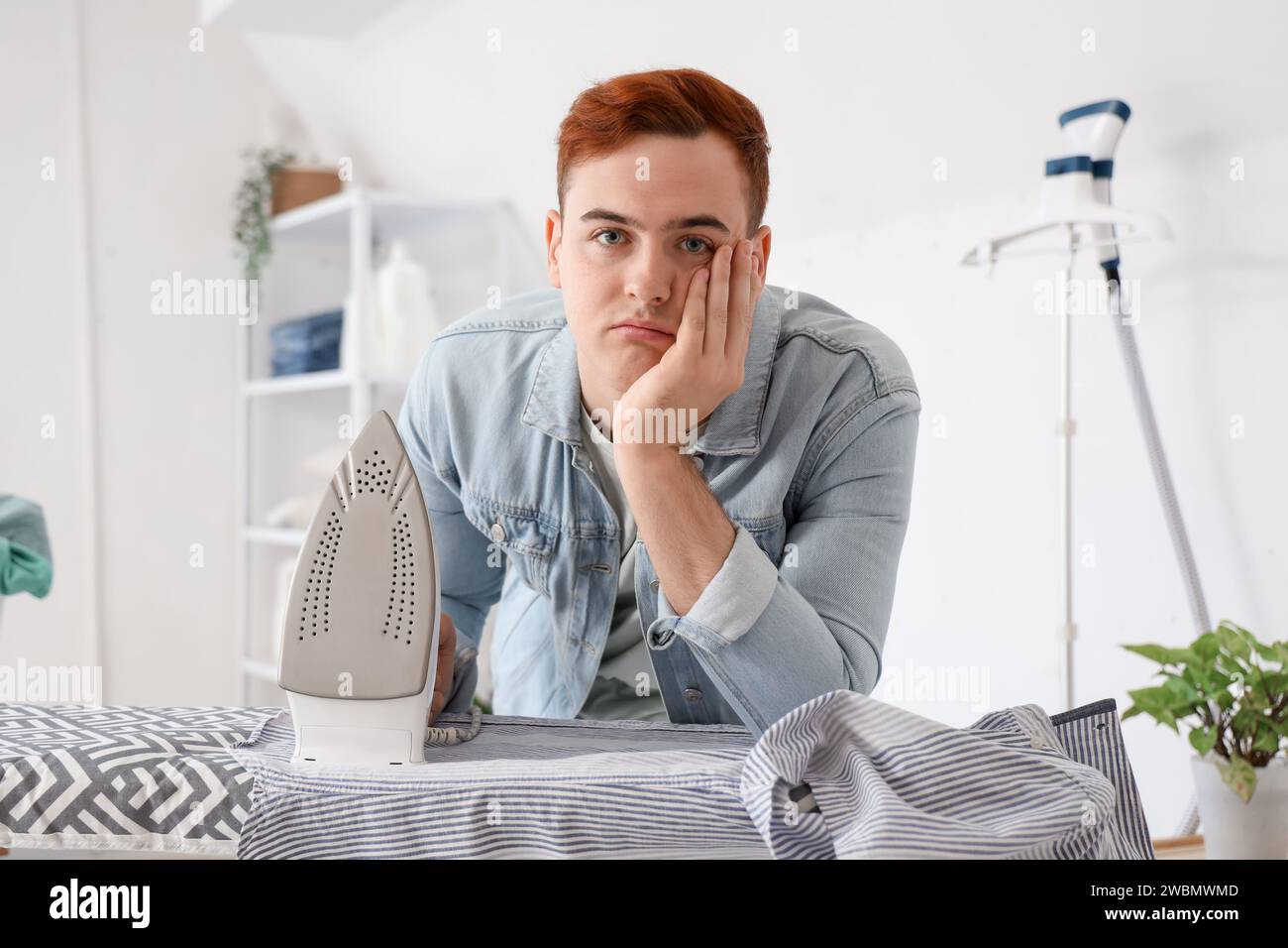 Bored young man with electric iron leaning on ironing board in laundry ...
