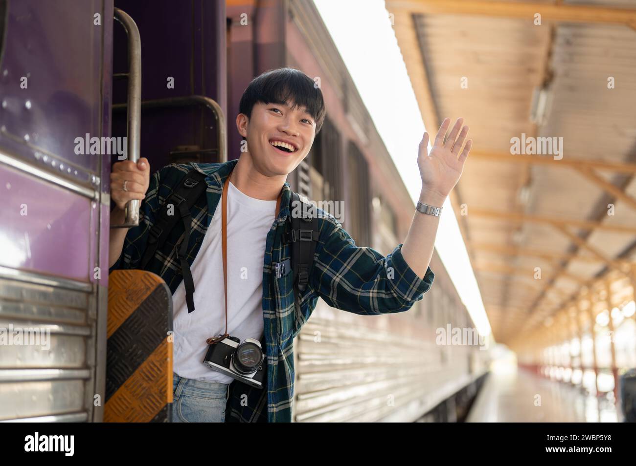 A cheerful young Asian male traveler is leaning out of the train door ...
