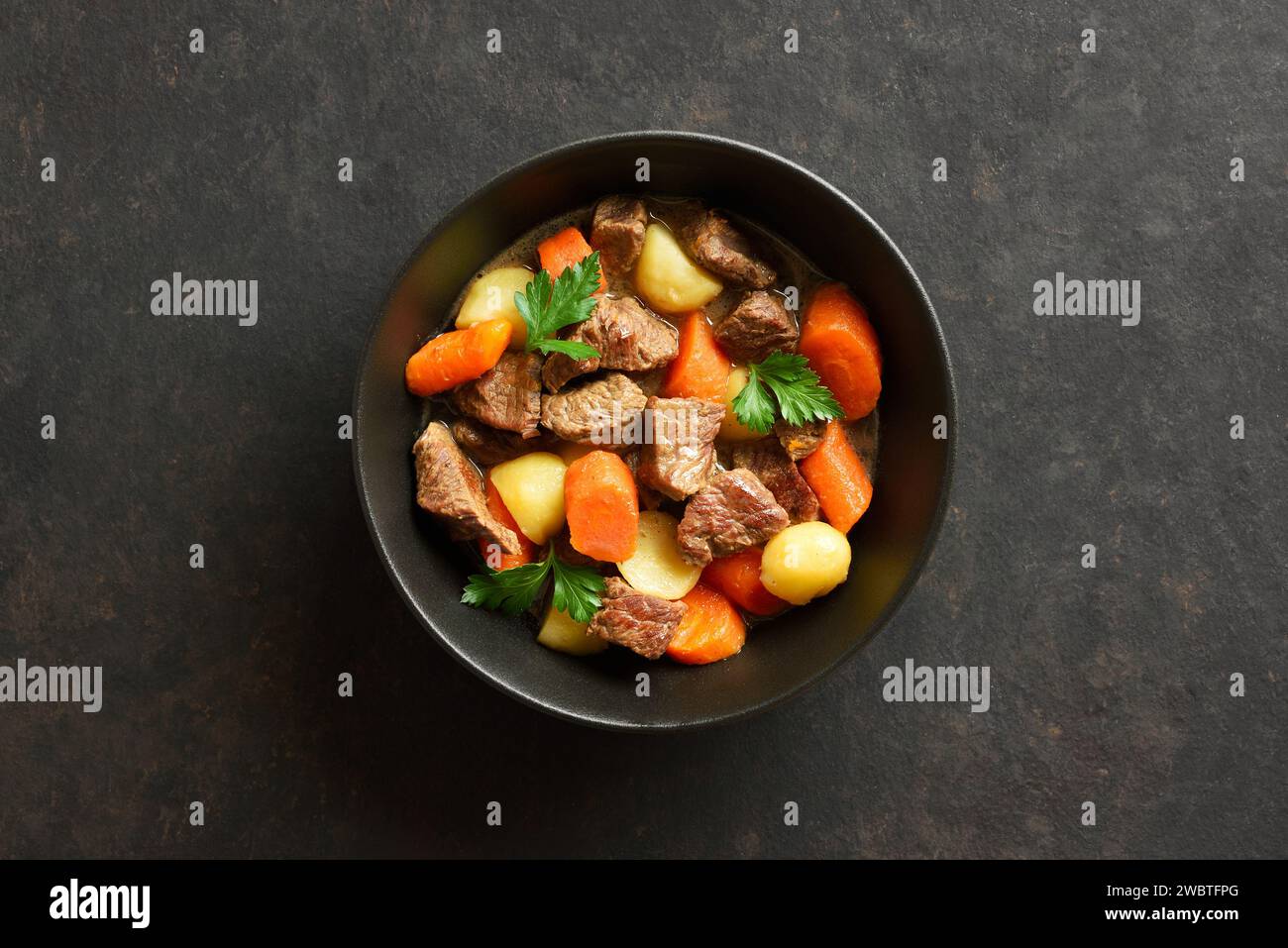 Beef stew with potatoes, carrots and greens in black bowl over dark ...