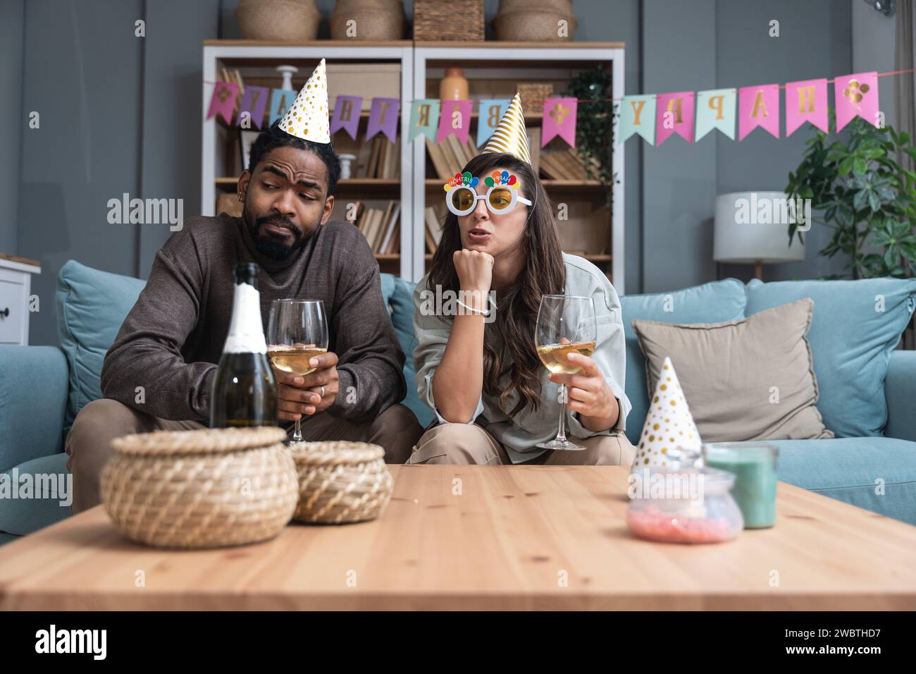 Two young people sitting on sofa with birthday caps, sad because no one ...