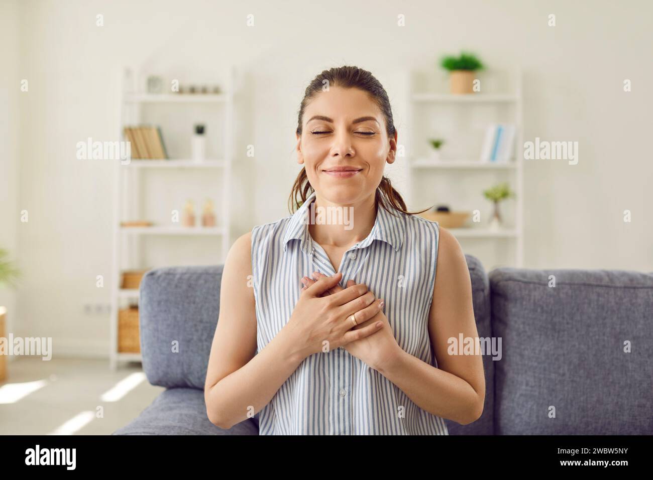 Woman sitting at home with her hands on chest with closed eyes and ...