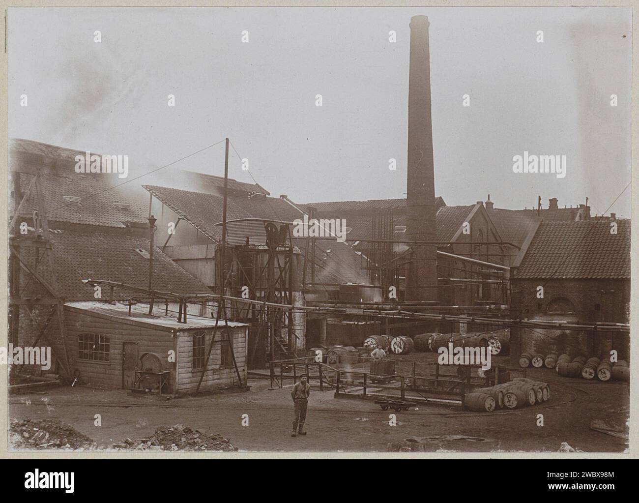 Factory site with buildings, a chimney, wooden barrels and an employee ...
