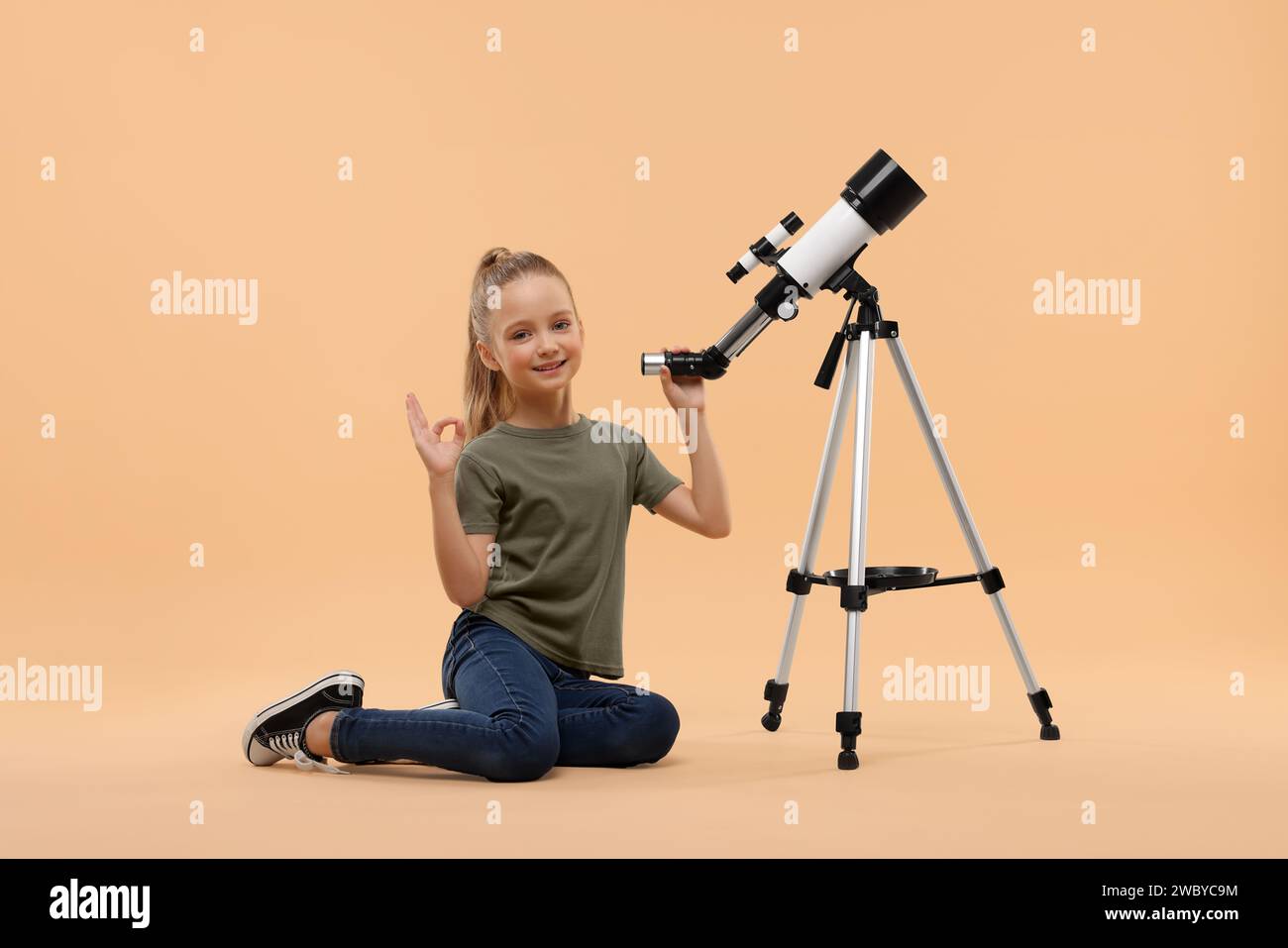 Happy little girl with telescope showing ok gesture on beige background ...