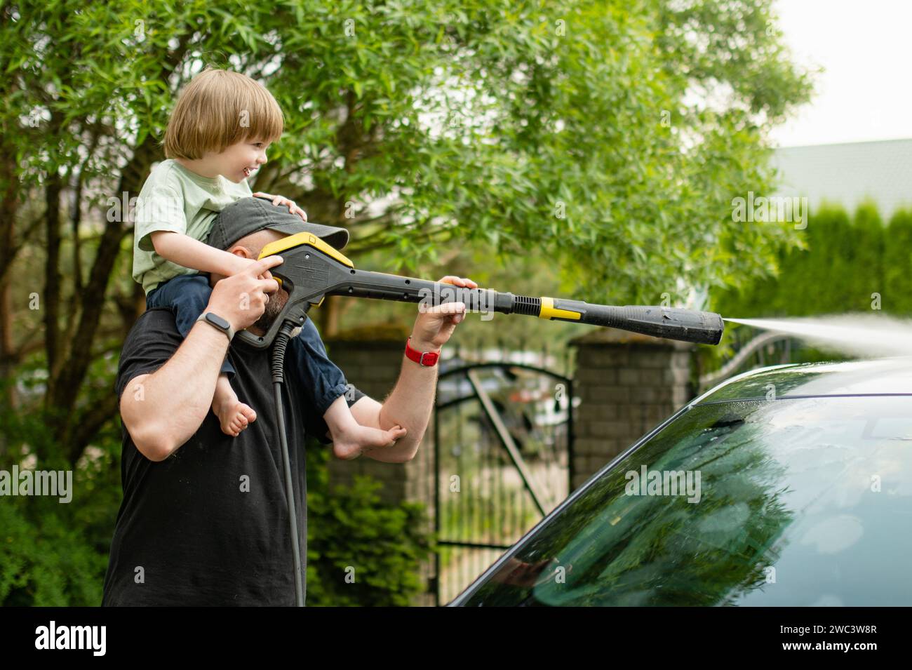 Father and his toddler son using a water gun to wash a car. Male driver ...