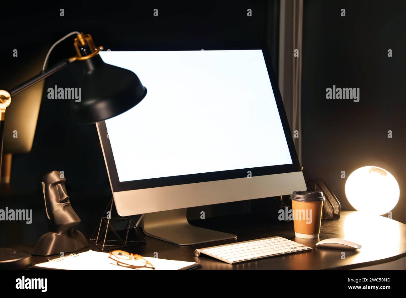 Blank computer monitor on table and glowing lamps in office at night ...